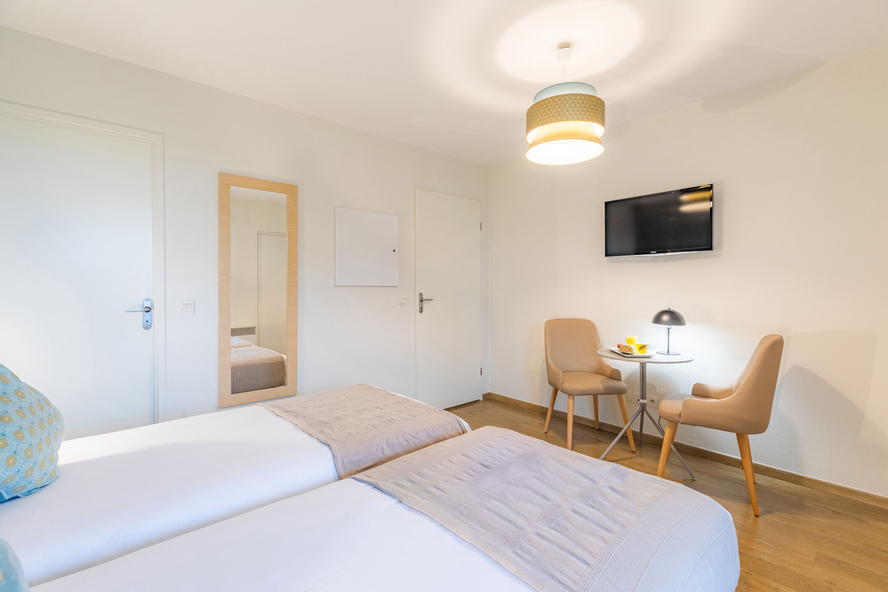Quarto de hotel com cama, escrivaninha com duas cadeiras, televisão na parede e luminária no teto.