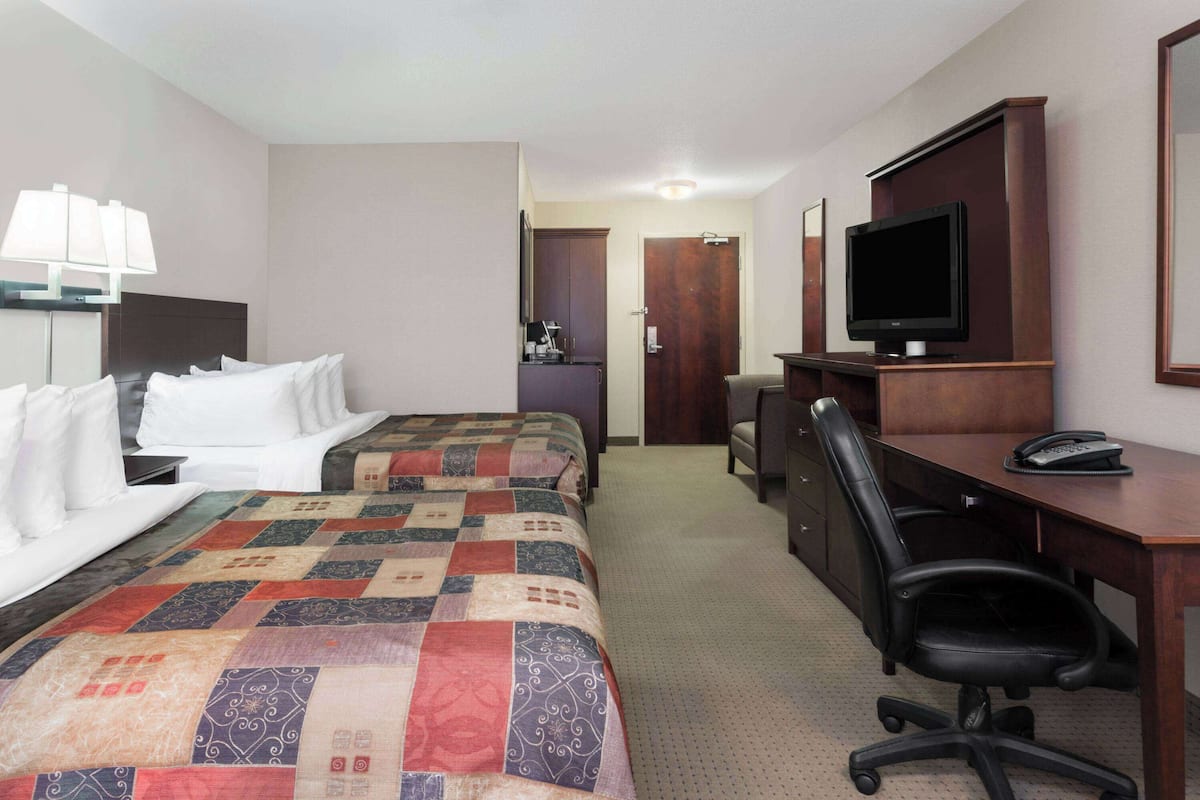 Quarto de hotel com duas camas, uma escrivaninha, uma cadeira, uma televisão e uma porta que dá para outro cômodo.