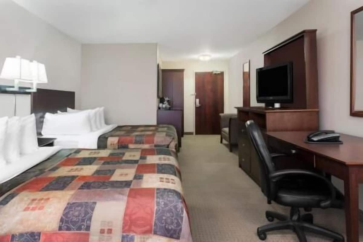 Quarto de hotel com duas camas, uma escrivaninha, uma cadeira, uma televisão e uma porta que dá para outro cômodo.