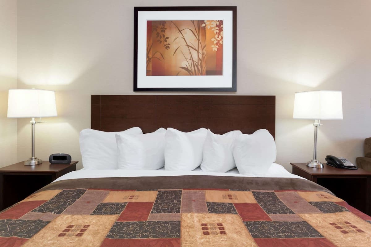 Quarto de hotel com uma cama grande, almofadas brancas, um edredom, duas mesas de cabeceira com abajures, um quadro na parede e uma cabeceira de madeira.
