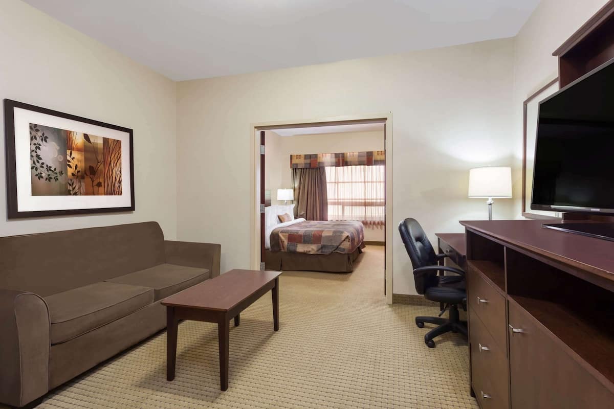 Quarto de hotel com sofá marrom, mesa de madeira, televisão em um armário de madeira, cama com manta colorida e quadro pendurado na parede.