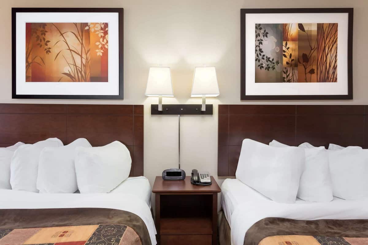Quarto de hotel com duas camas, uma mesa de cabeceira com telefone e relógio, e duas luminárias fixadas na parede.