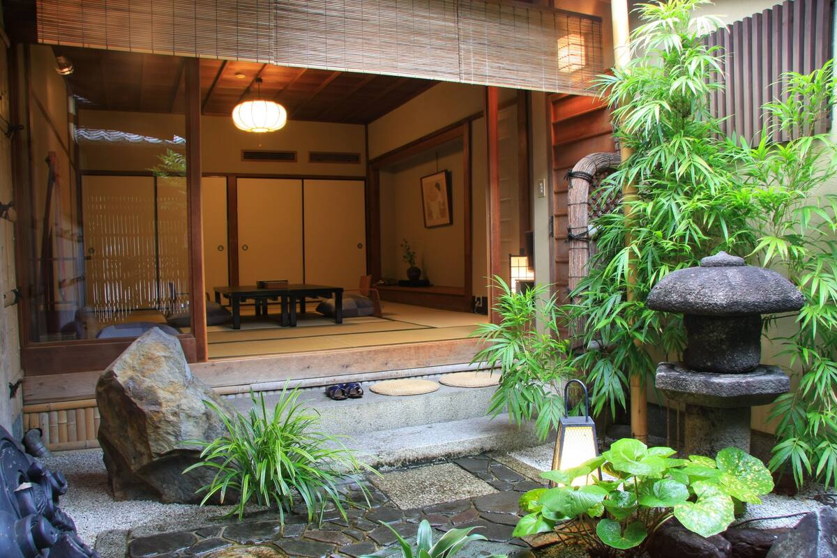 Um interior japonês tradicional com tatames, um lampião de pedra e uma escadaria de madeira que leva a um quarto.