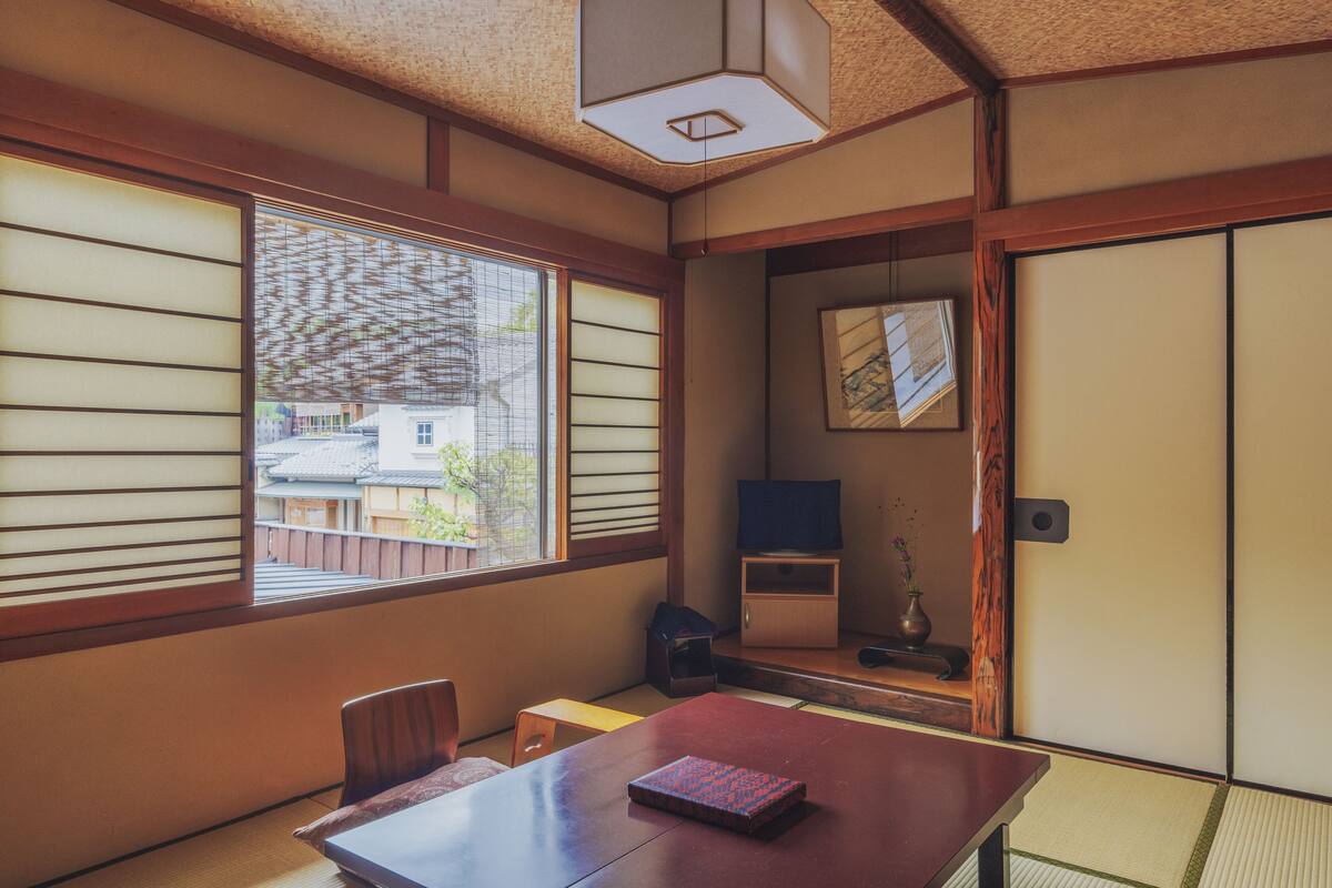 Um quarto japonês tradicional com tatames, uma mesa de madeira baixa, uma televisão e portas de correr.