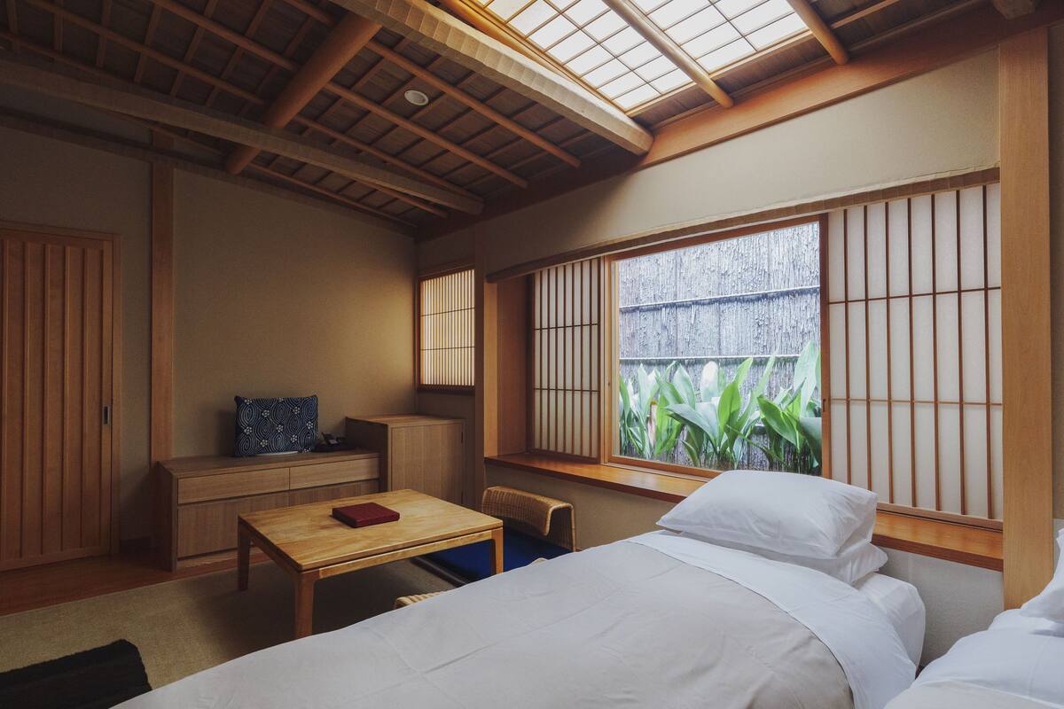 Um quarto japonês tradicional com piso de tatame, uma mesa de madeira baixa, uma cama com roupa de cama branca e portas deslizantes shoji.