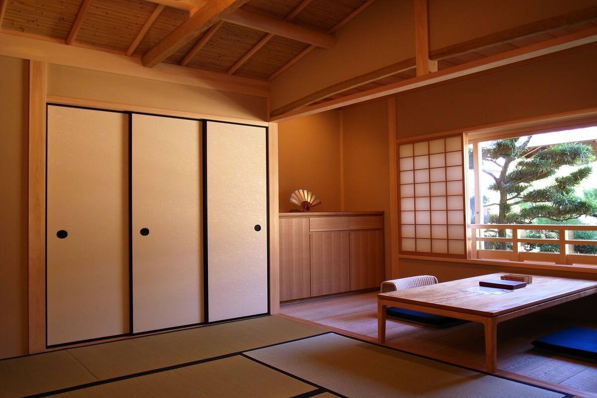 Um quarto japonês tradicional com tatames, portas deslizantes, uma mesa baixa e vista para uma árvore através de um painel shoji.