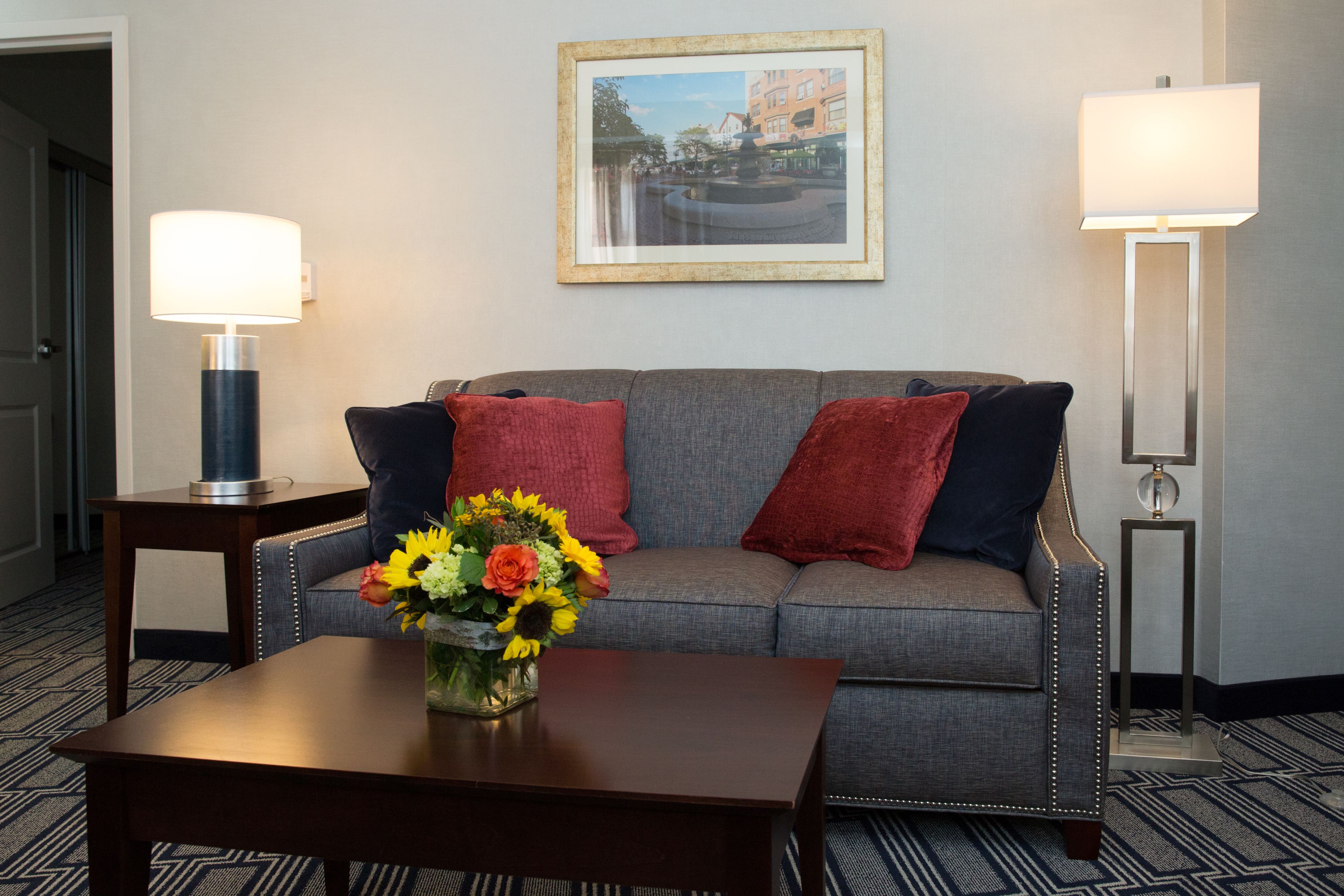 Quarto de hotel com sofá cinza, duas almofadas vermelhas, um vaso com flores, uma mesa de centro de madeira e duas luminárias.