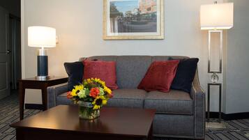 Quarto de hotel com sofá cinza, duas almofadas vermelhas, um vaso com flores, uma mesa de centro de madeira e duas luminárias.