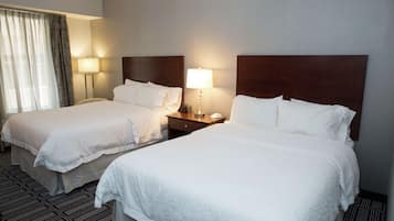 Quarto de hotel com duas camas, cada uma com roupa de cama branca, mesa de cabeceira com abajur e janela com cortinas.