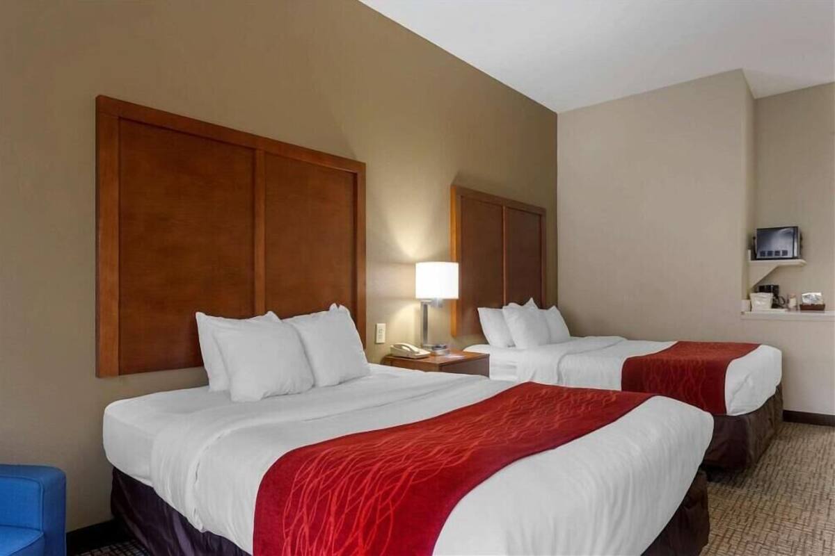 Quarto de hotel com duas camas, uma cabeceira de madeira, uma mesa de cabeceira com abajur e uma televisão na parede.