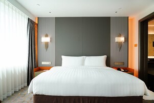 Quarto de hotel com uma cama grande, duas mesas de cabeceira, um criado-mudo, uma luminária na parede e uma janela com cortinas.