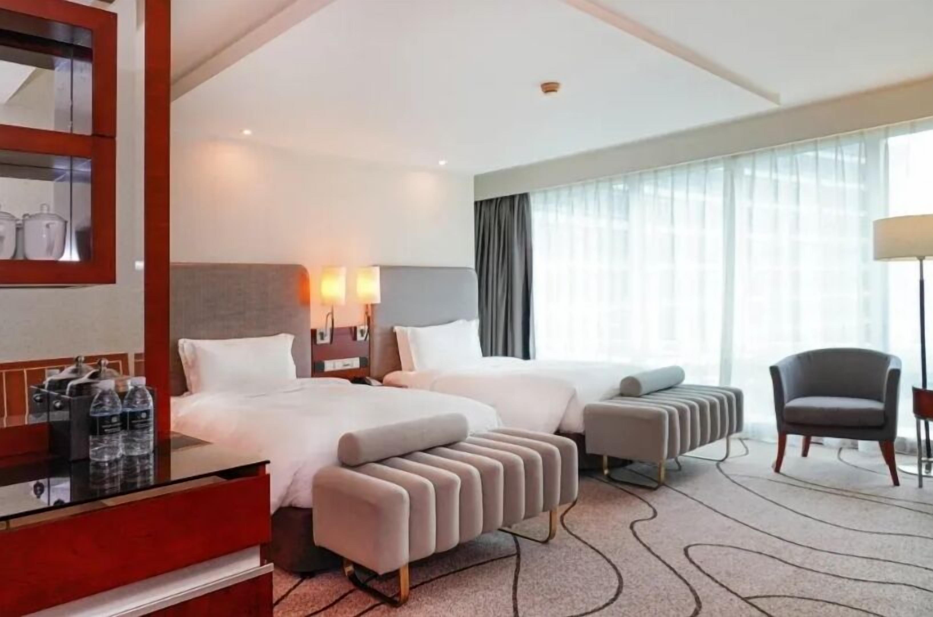 Quarto de hotel com duas camas, uma escrivaninha, uma cadeira e uma janela grande com cortinas.