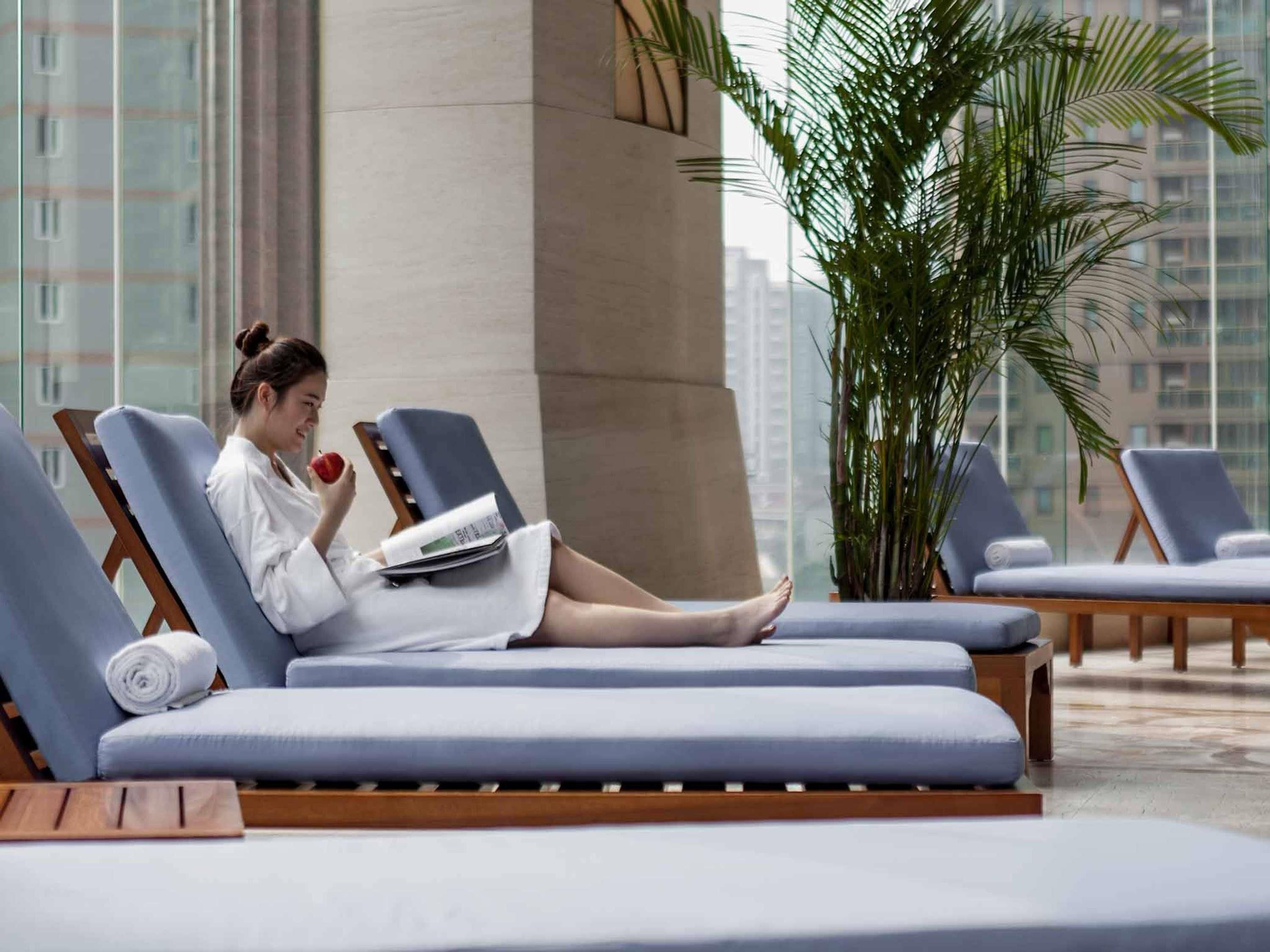 Uma pessoa relaxando em uma espreguiçadeira com um livro e um objeto vermelho, em um ambiente interno moderno com uma planta grande e vista para edifícios.