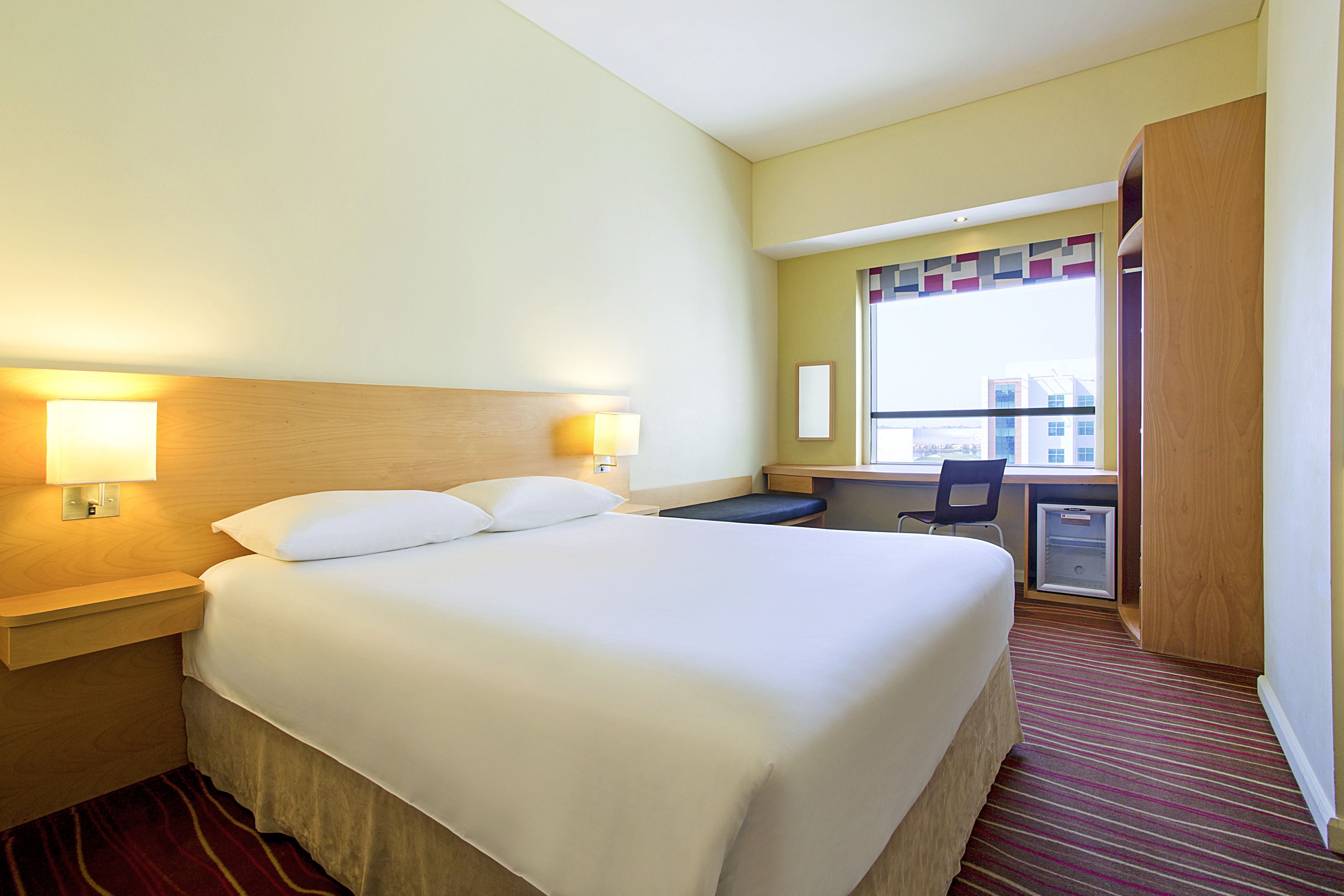 Quarto de hotel com uma cama grande, duas luminárias de cabeceira, uma escrivaninha com cadeira e vista para a cidade pela janela.