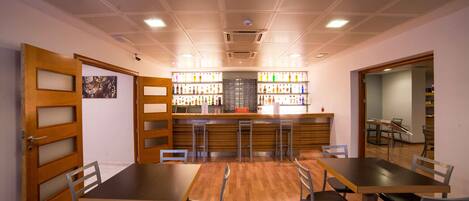 Uma sala de conferências com uma mesa comprida, cadeiras, área de bar com prateleiras de garrafas e uma porta de madeira com painéis de vidro jateado.
