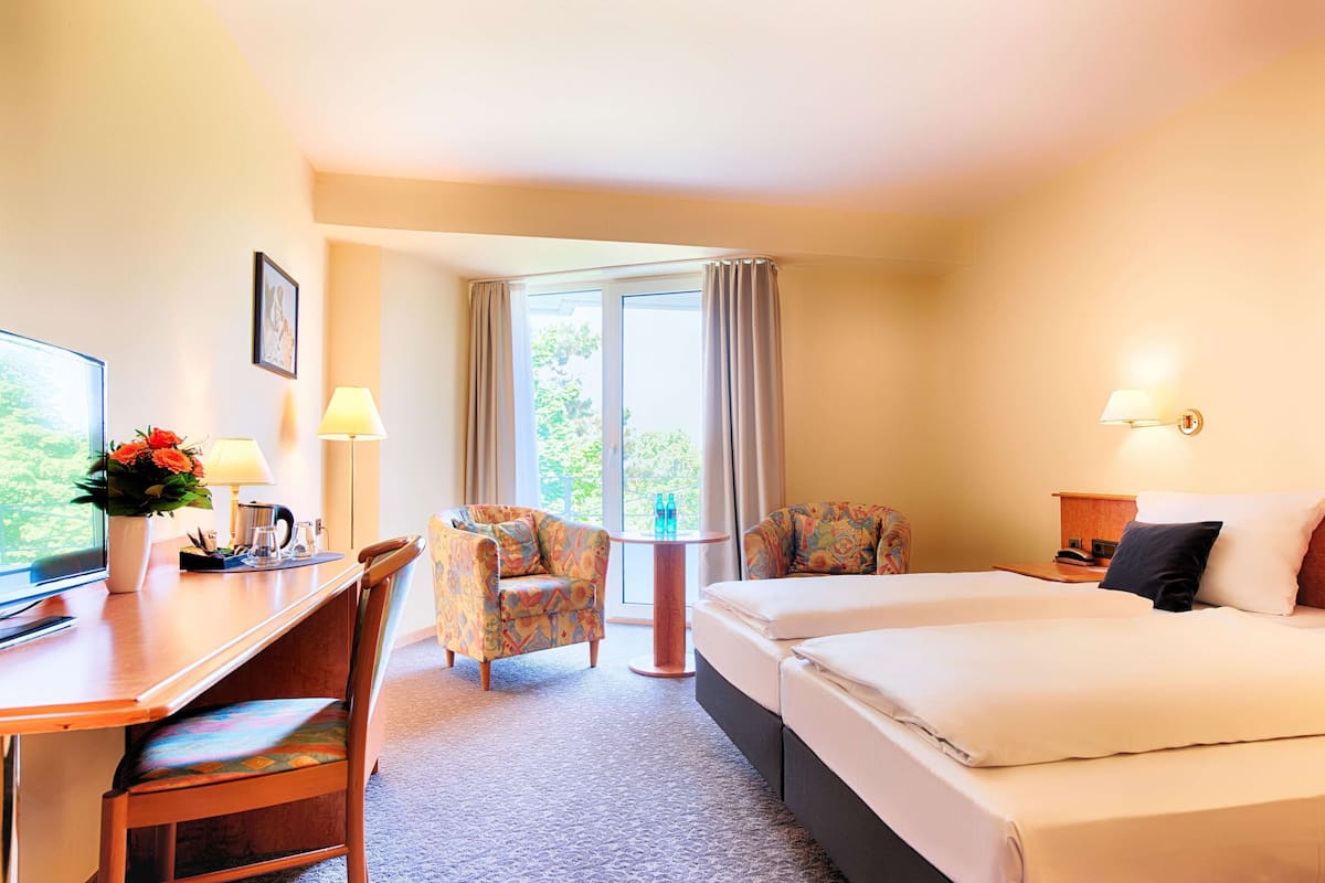 Quarto de hotel com cama, escrivaninha com computador, cadeira, mesinha com abajur e janela com cortinas.