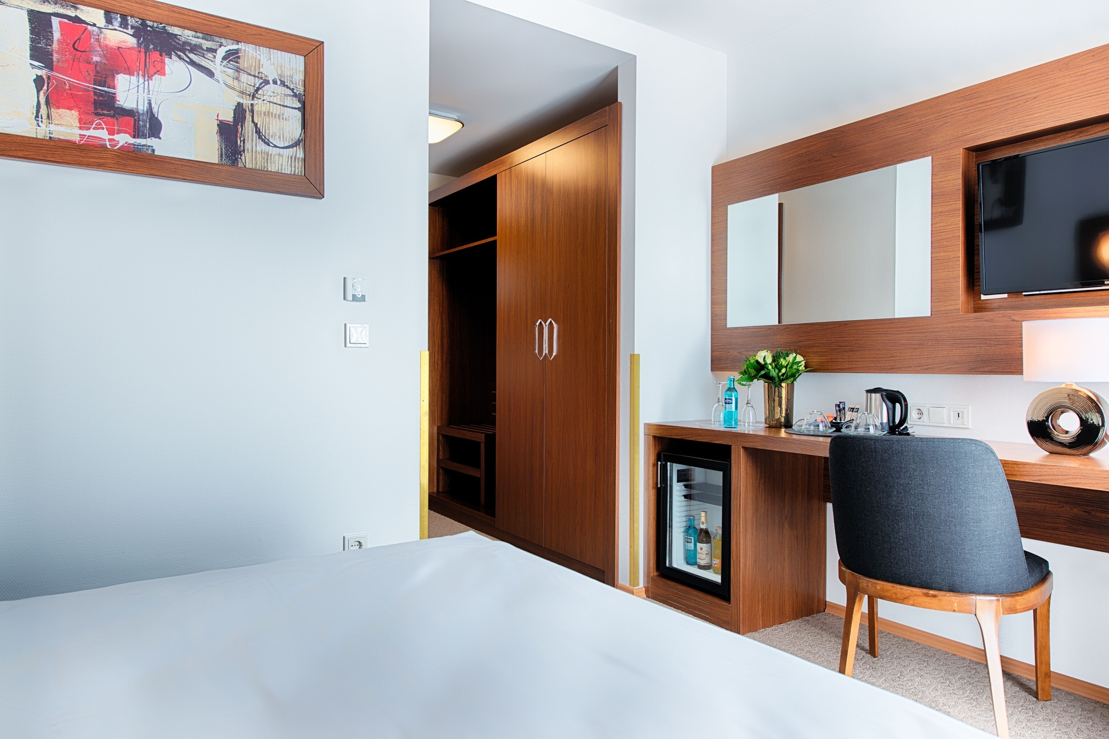 Quarto de hotel moderno com escrivaninha de madeira, uma cadeira, minibar e um espelho grande.