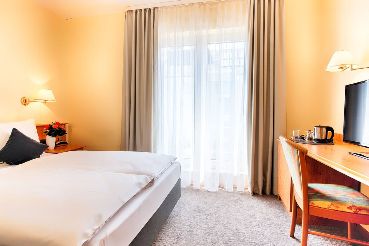 Quarto de hotel com cama, escrivaninha com cadeira, televisão e janela com cortinas.