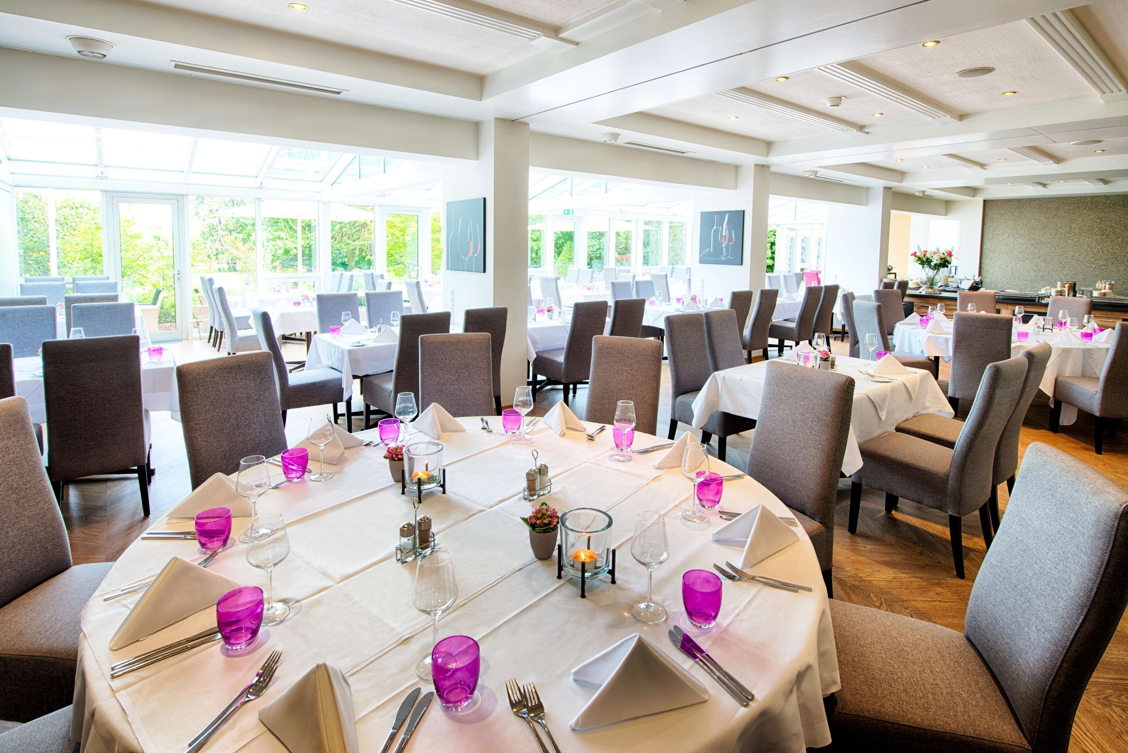 Uma sala de jantar ampla com mesas redondas dispostas para um evento formal, com toalhas de mesa brancas e copos de vidro roxos.