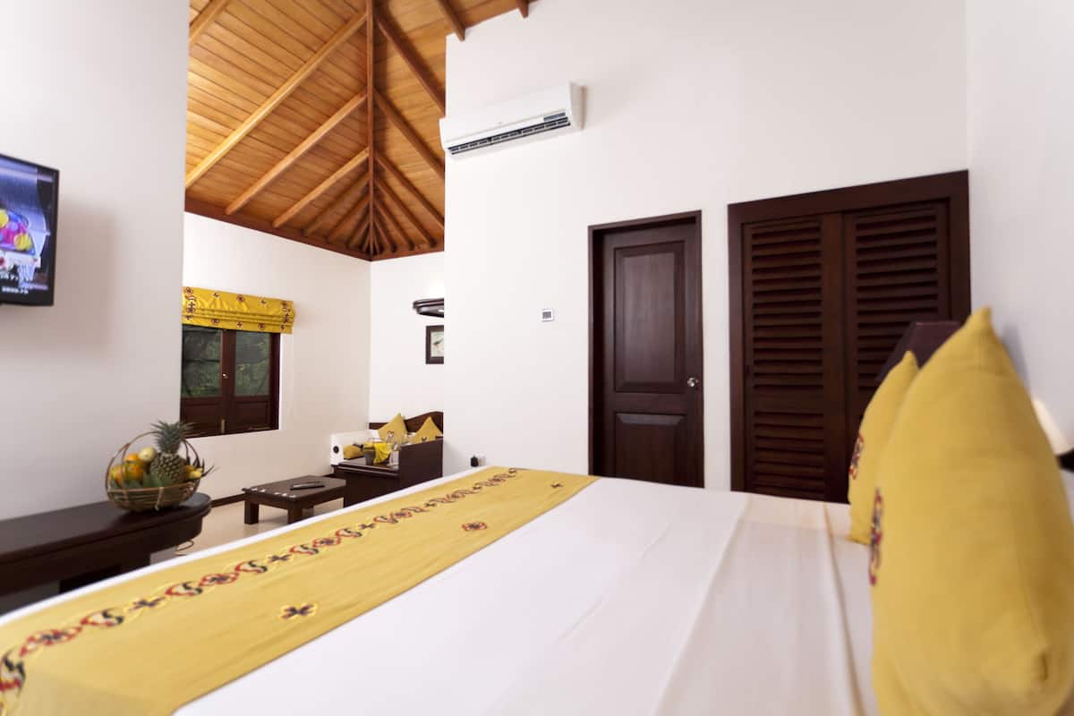 Quarto de hotel com uma cama, televisão, teto de madeira e janela com cortinas amarelas.