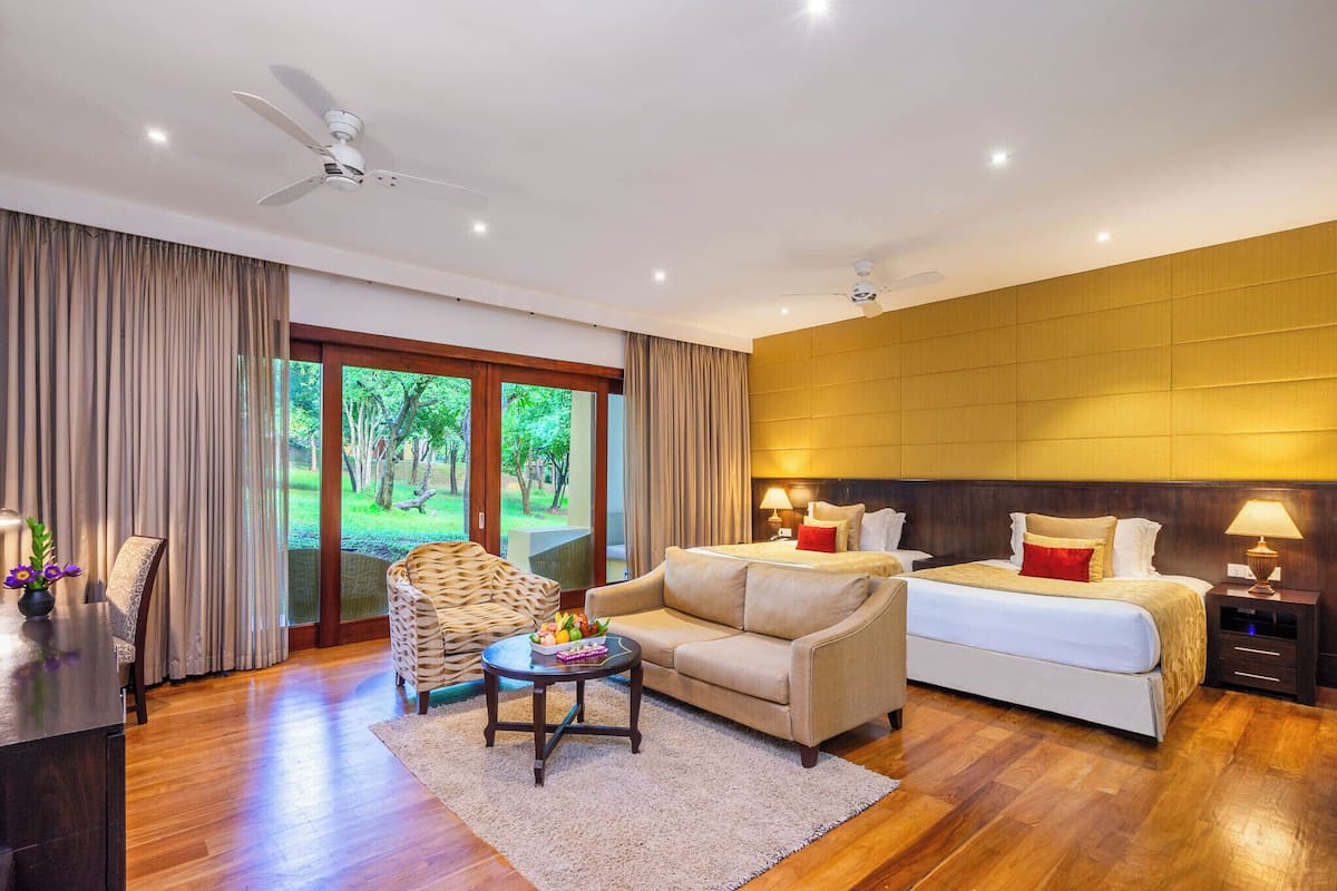 Quarto de hotel com cama, sofá, poltrona, mesa pequena e escrivaninha. Há uma janela com cortinas e vista para o verde lá fora.