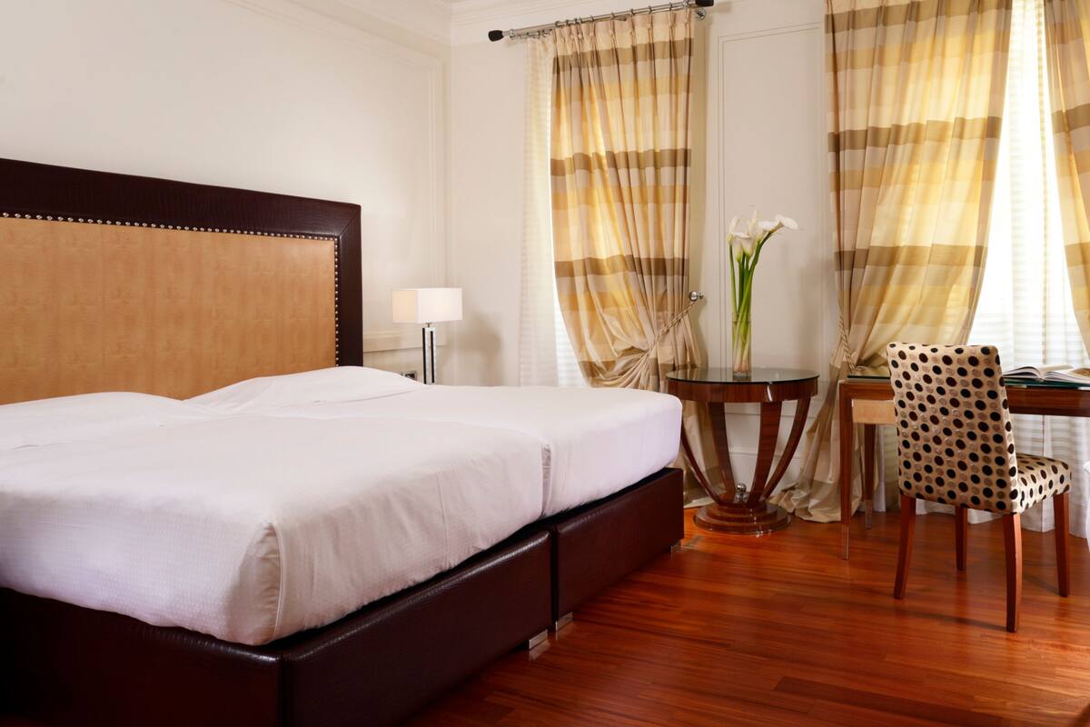 Quarto de hotel com uma cama grande, uma mesinha com um vaso de flores e uma cadeira estampada em bolinhas.