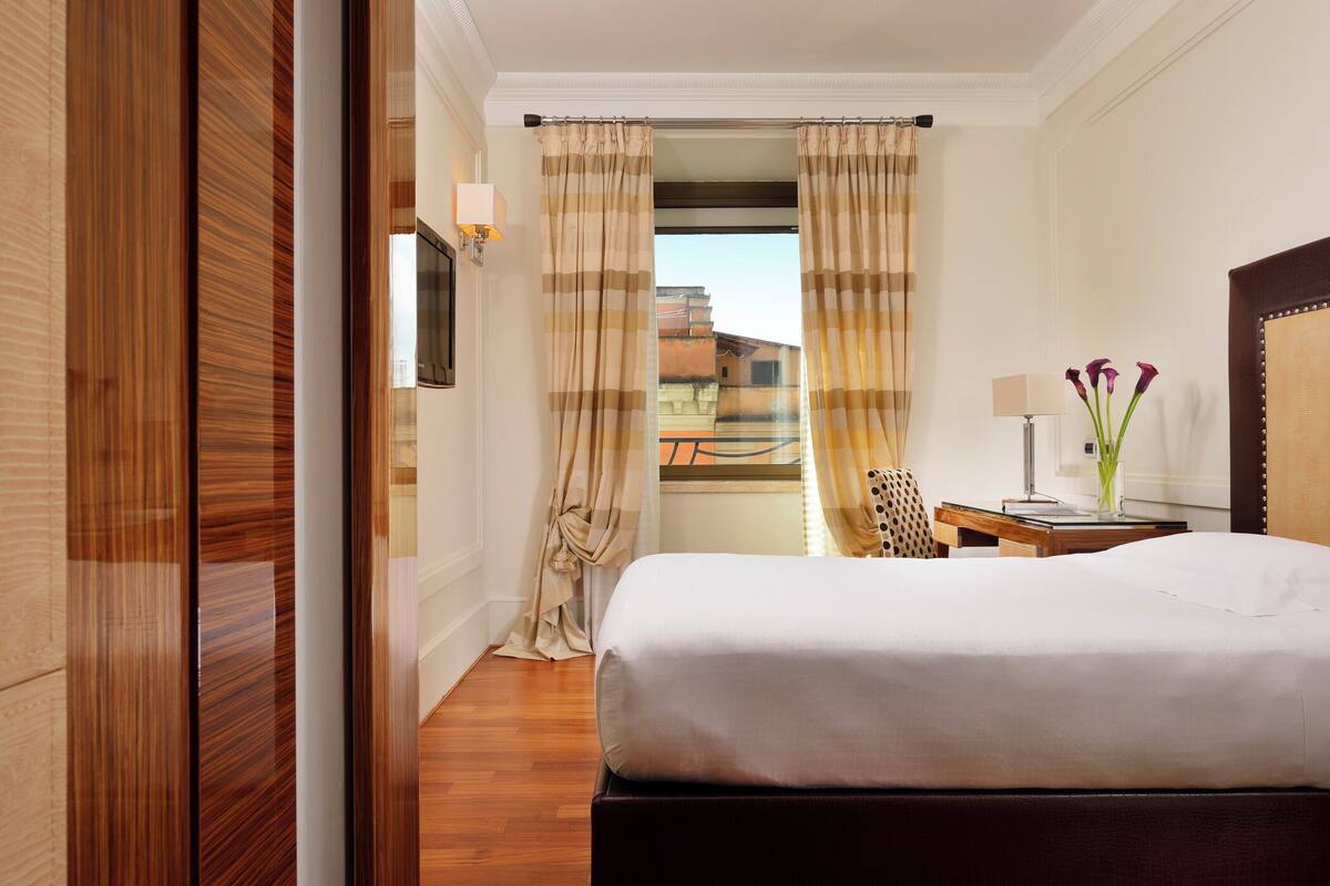 Quarto de hotel com uma cama, uma escrivaninha, uma cadeira, um abajur e vista para edifícios através de cortinas.