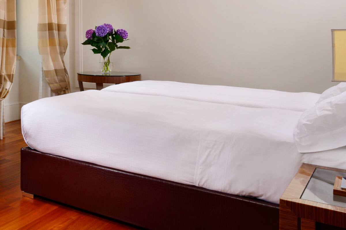 Quarto de hotel com uma cama grande, piso de madeira, uma mesinha de cabeceira com um vaso de flores e uma janela com cortinas.