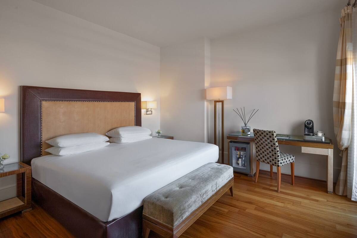 Quarto de hotel com uma cama grande, uma escrivaninha, uma cadeira e uma mesa de cabeceira com luminária.
