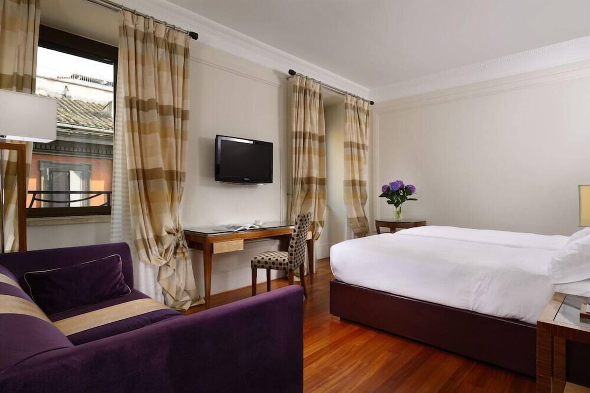 Quarto de hotel com cama, escrivaninha, cadeira, televisão e janela com cortinas.