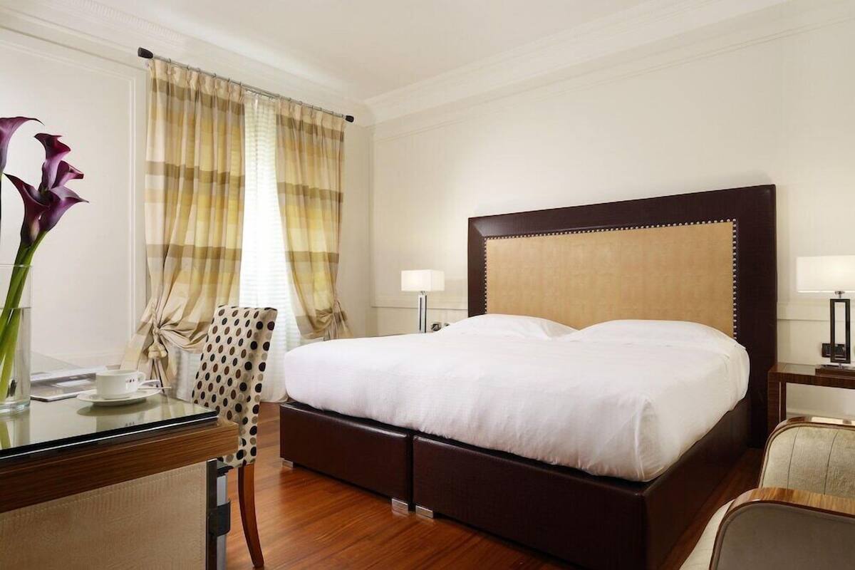 Quarto de hotel com uma cama grande, uma mesa com cadeira, um vaso com flores e uma janela com cortinas.