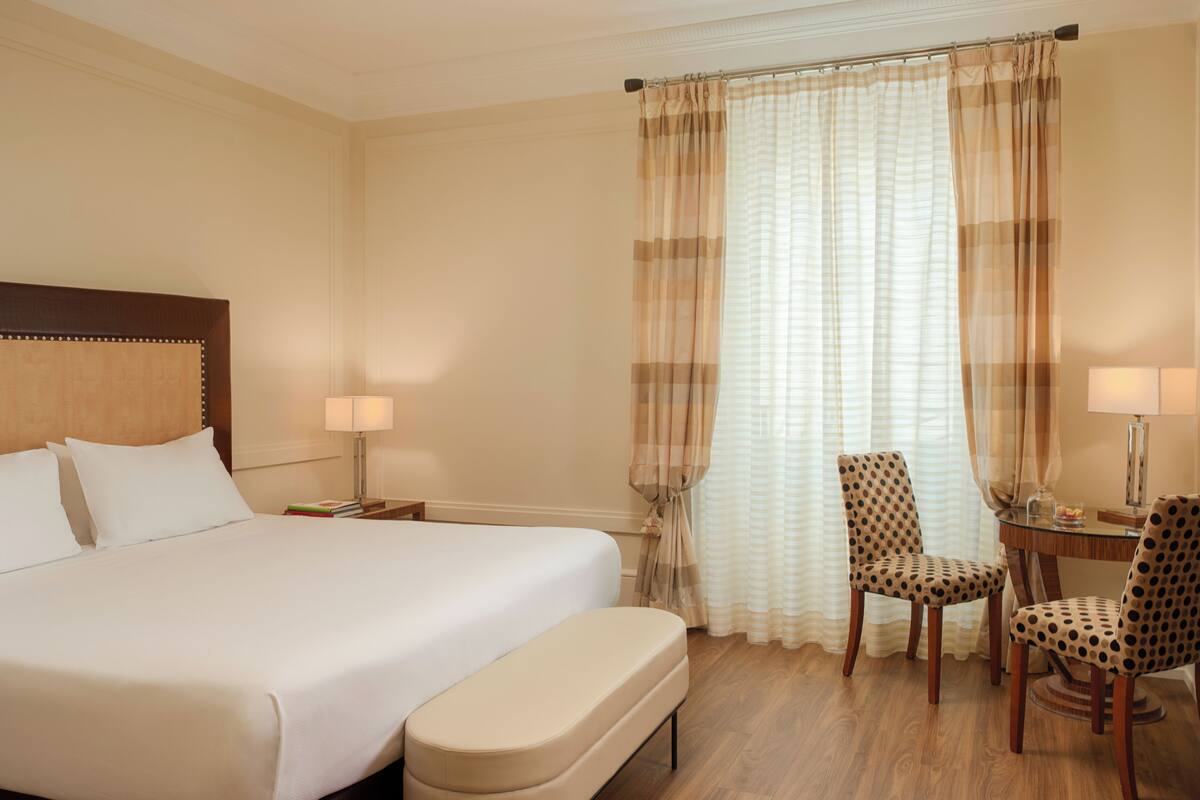 Quarto de hotel com uma cama grande, mesa de cabeceira, cadeira, escrivaninha e janela com cortinas.