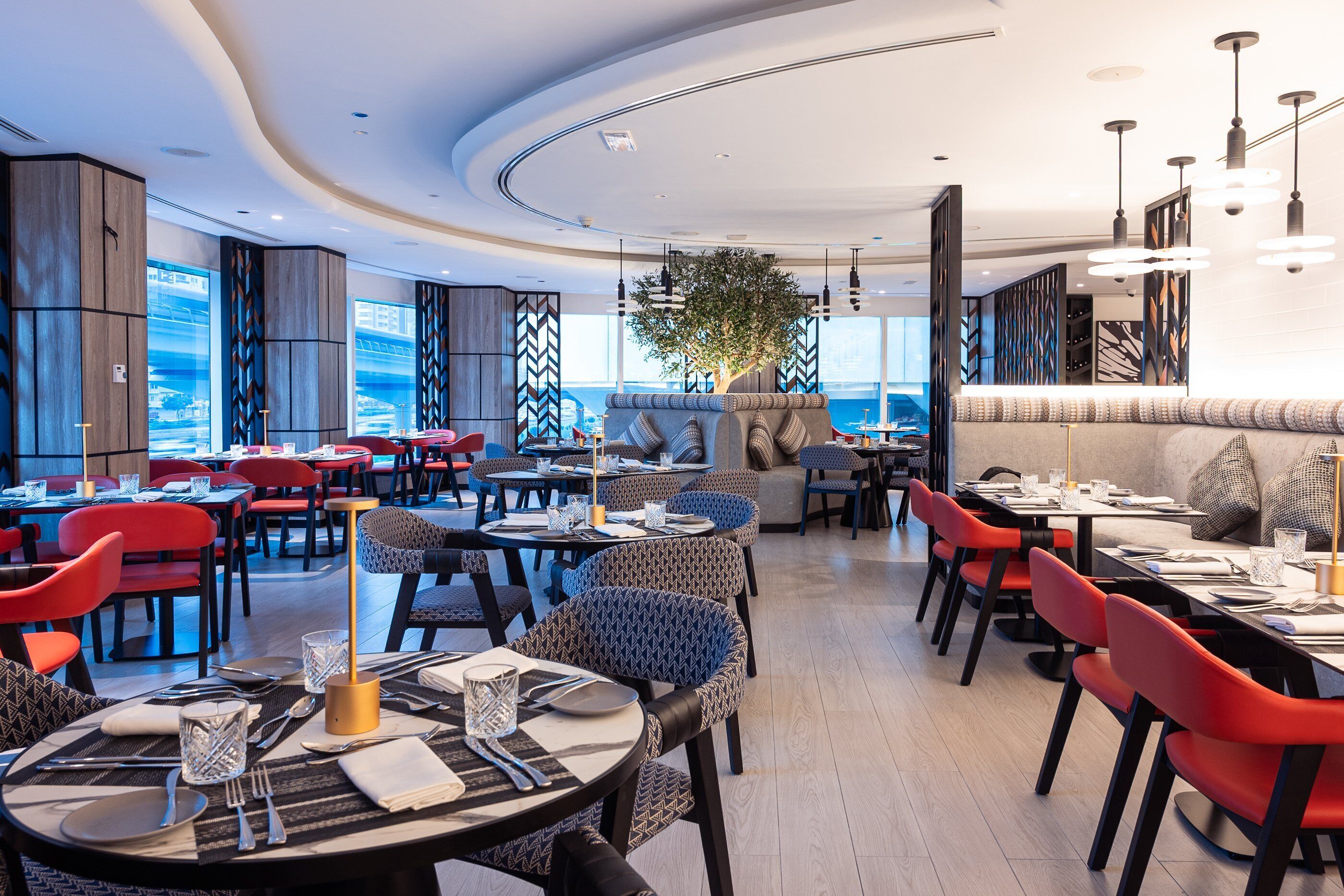 Restaurante moderno com mesas redondas, cadeiras vermelhas e cinzentas e uma grande árvore como peça central.