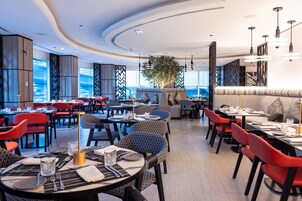Restaurante moderno com mesas redondas, cadeiras vermelhas e cinzentas e uma grande árvore como peça central.