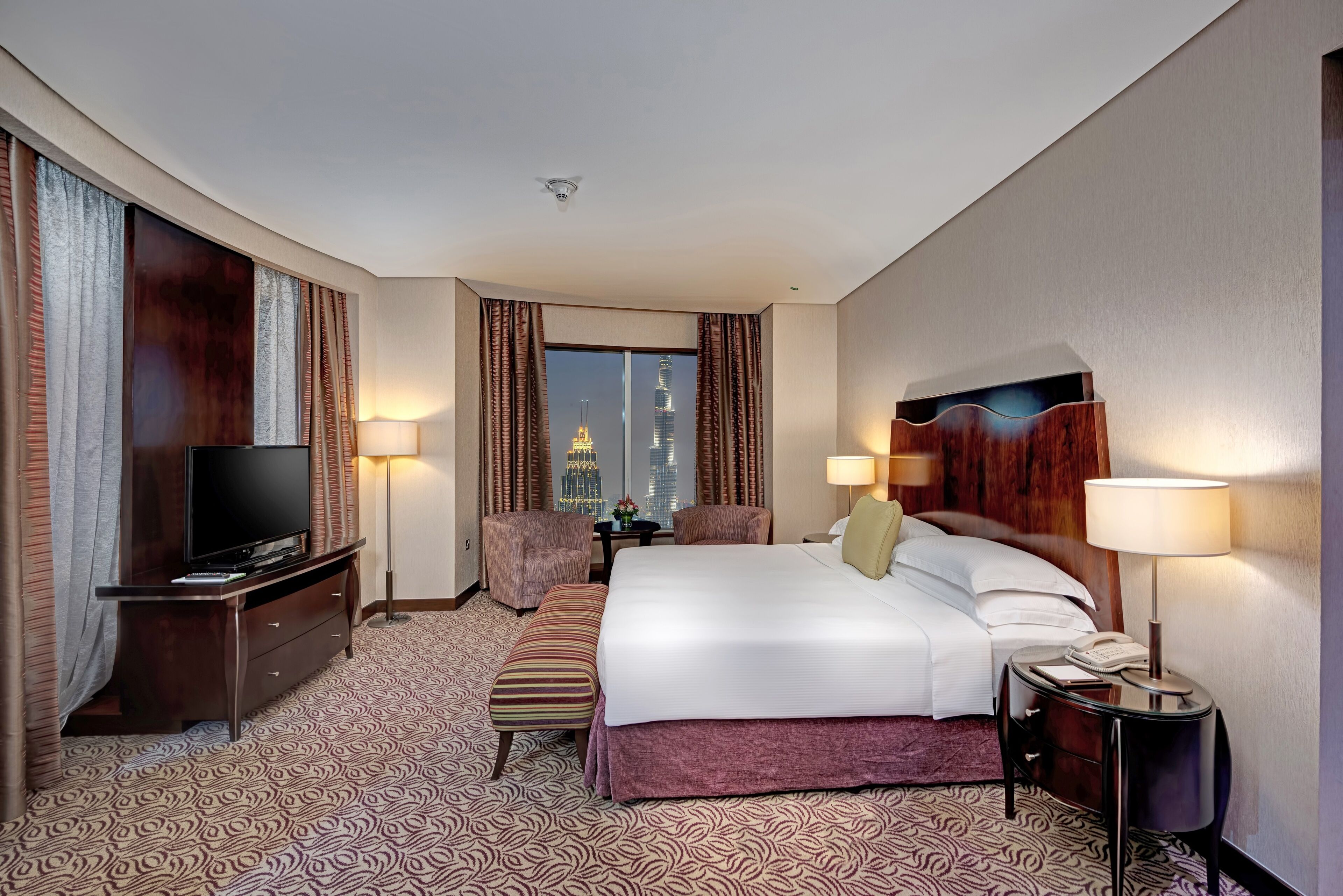 Quarto de hotel com uma cama grande, televisão, escrivaninha e vista para um prédio alto.