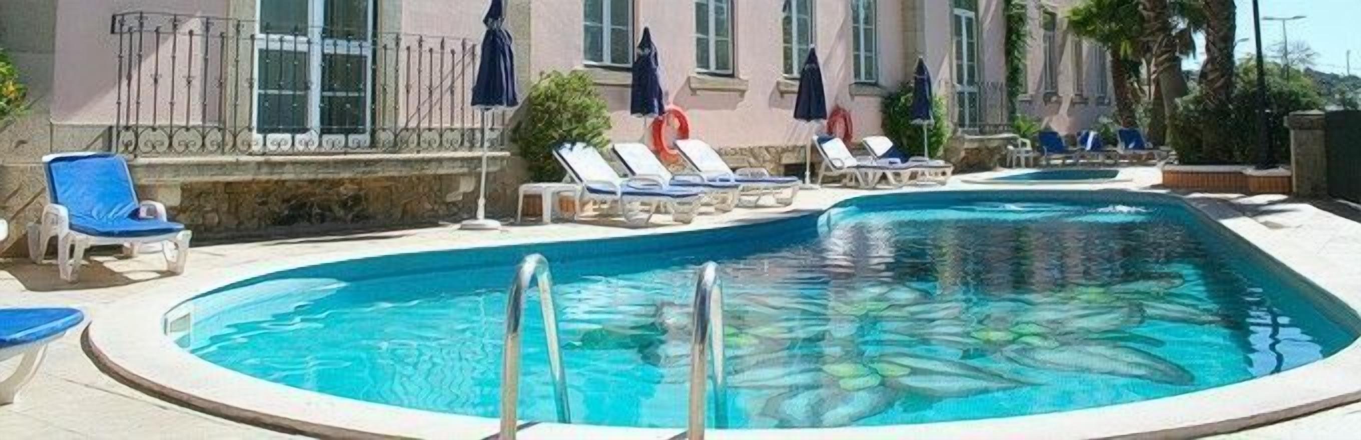 Uma piscina com espreguiçadeiras e guarda-sóis, rodeada por um edifício com fachada de pedra.
