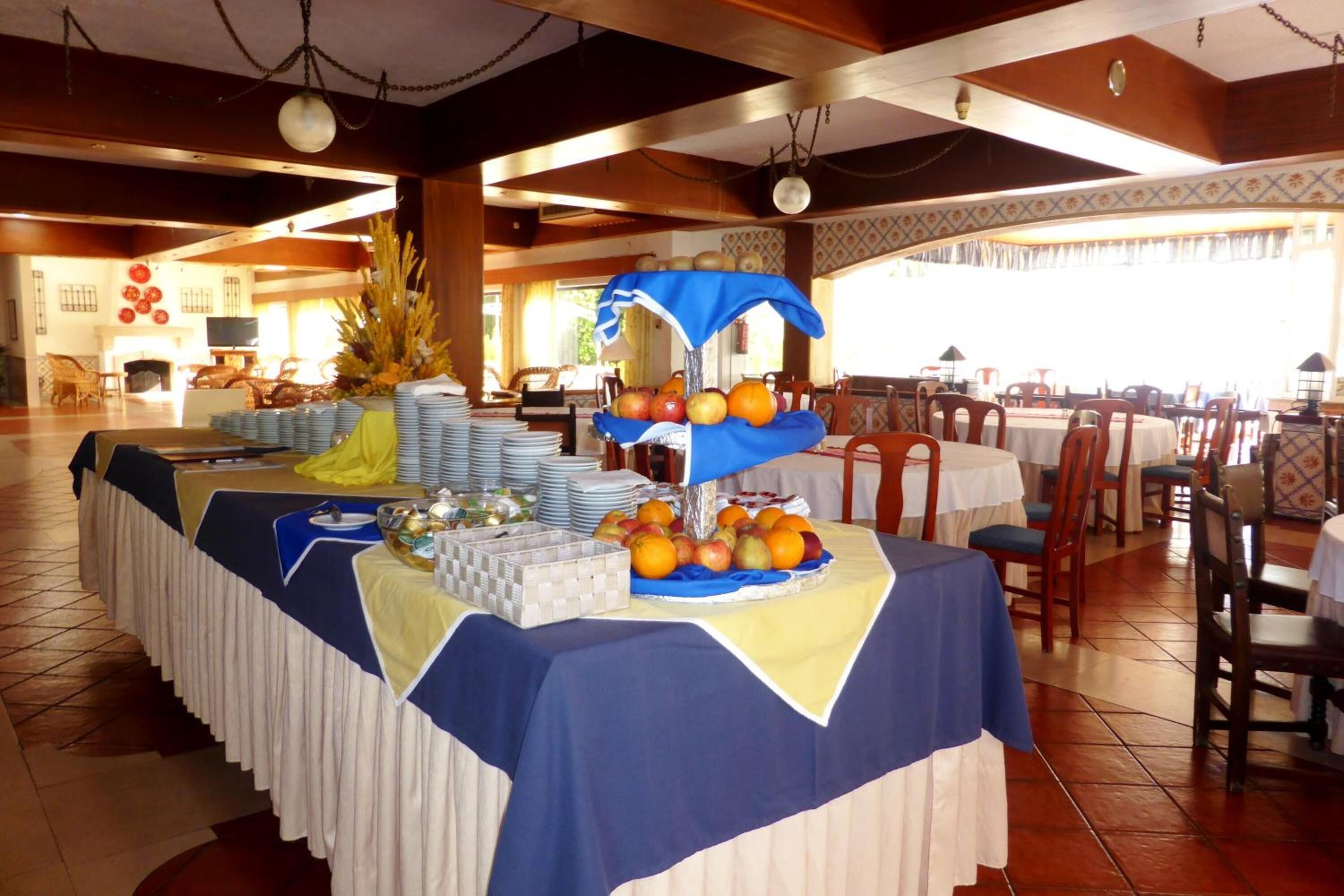 Disposição de buffet com várias mesas, cada uma coberta por toalhas azuis e brancas. Frutas e diversos pratos são exibidos nas mesas.