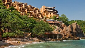 Um hotel à beira-mar com várias varandas, uma ponte de acesso à praia e vegetação exuberante.