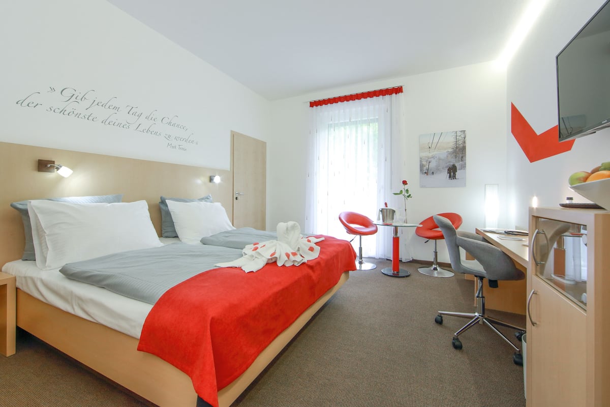 Quarto de hotel com cama, escrivaninha, cadeira, televisão e parede com uma citação.