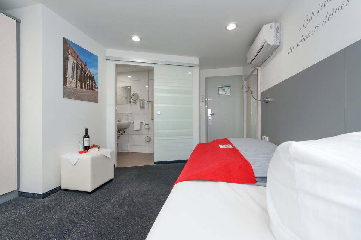 Quarto de hotel com uma cama, uma pequena mesa branca com uma garrafa e copos, um cobertor vermelho na cama, um quadro na parede e um banheiro visível por uma porta aberta.