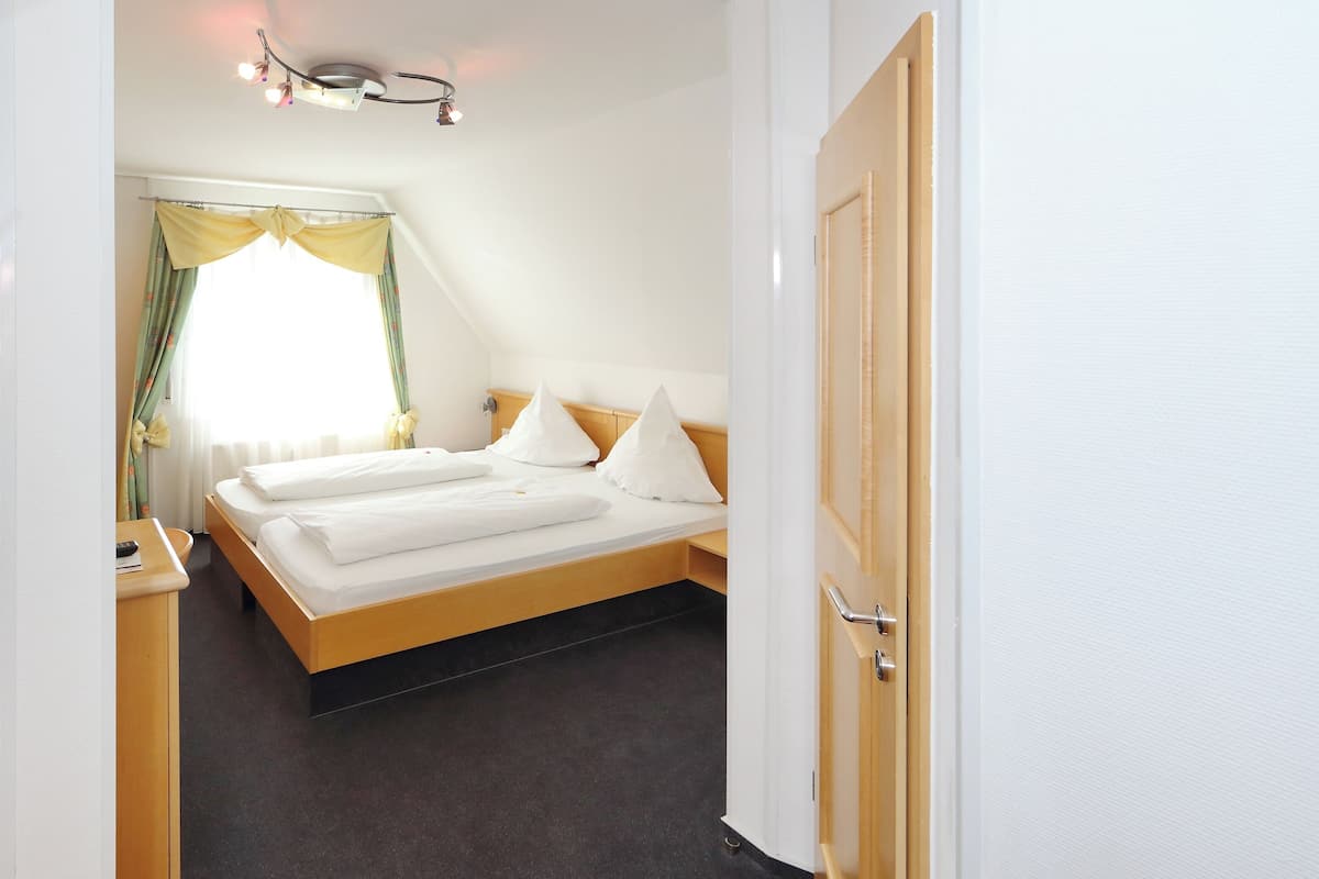 Quarto de hotel com uma cama de casal, mesa de cabeceira, escrivaninha e janela com cortinas.