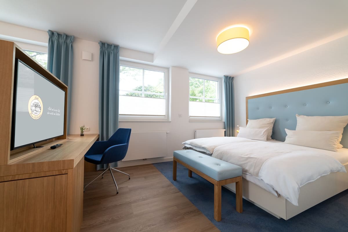 Quarto de hotel com uma cama, uma escrivaninha com computador, uma cadeira e uma janela com cortinas.