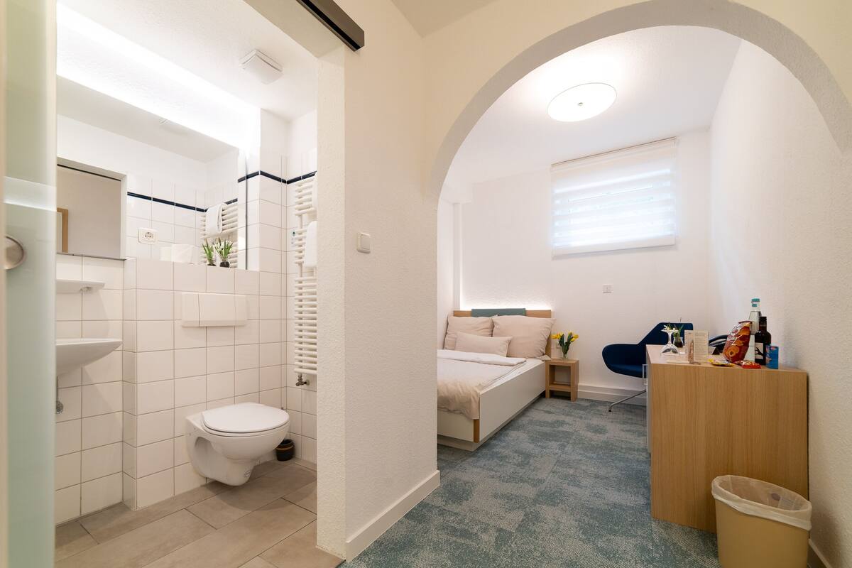 Uma casa de banho moderna com sanita, lavatório e chuveiro. Há uma cama com uma mesa de cabeceira e uma pequena mesa com uma lâmpada.