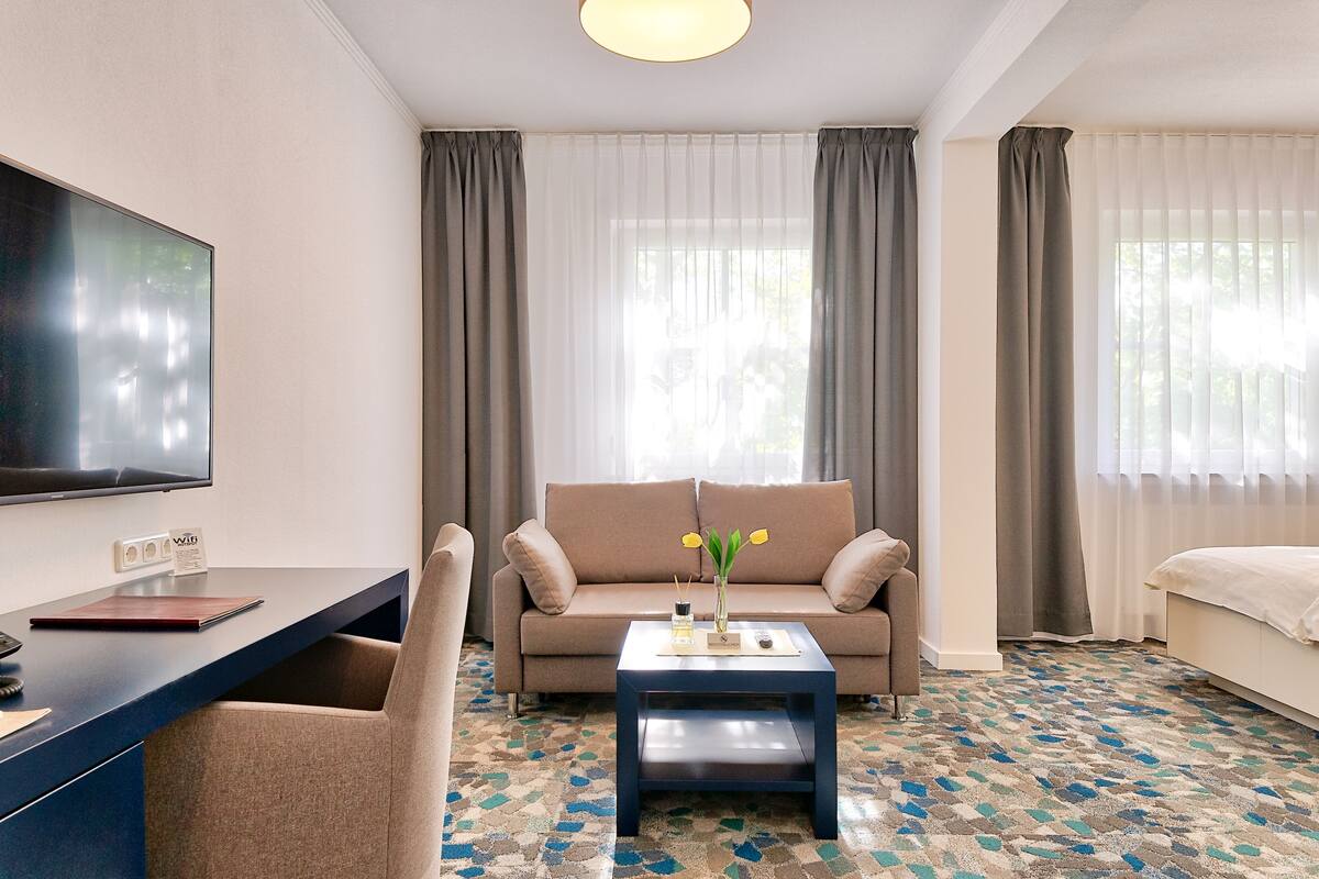 Quarto de hotel com sofá, mesa de centro com um vaso, televisão e carpete estampado.