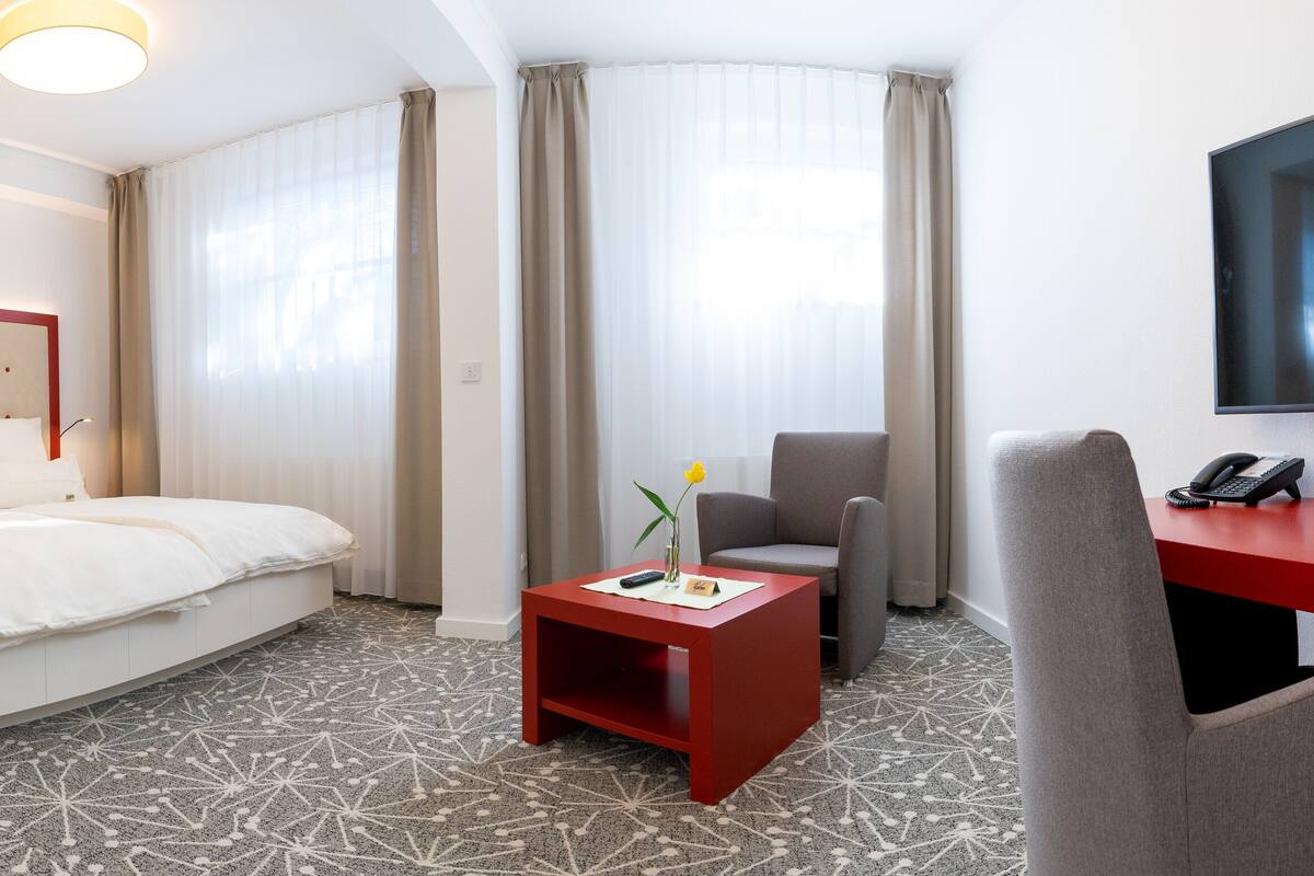 Quarto de hotel com cama, escrivaninha, cadeira, televisão e uma pequena mesa com um vaso.