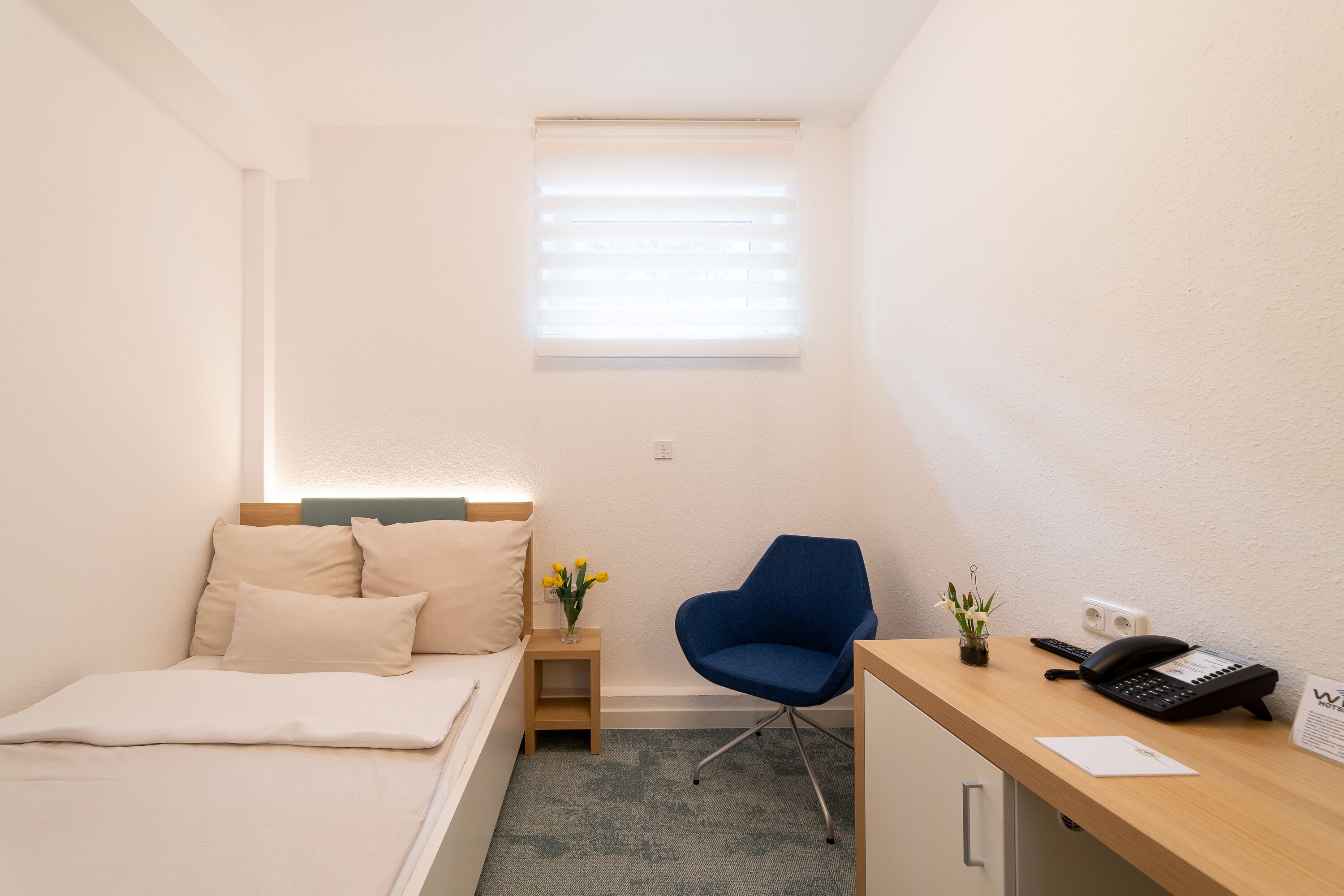 Quarto pequeno e bem organizado, com cama, escrivaninha, cadeira, mesinha de centro e um vaso de flores, além de uma janela com persianas.