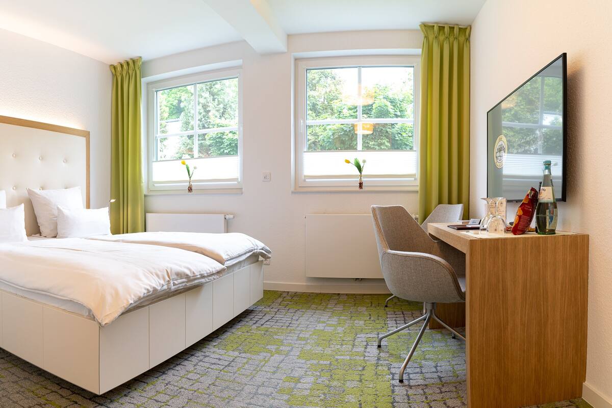 Quarto de hotel com cama, escrivaninha, cadeira, espelho grande e duas janelas com cortinas.