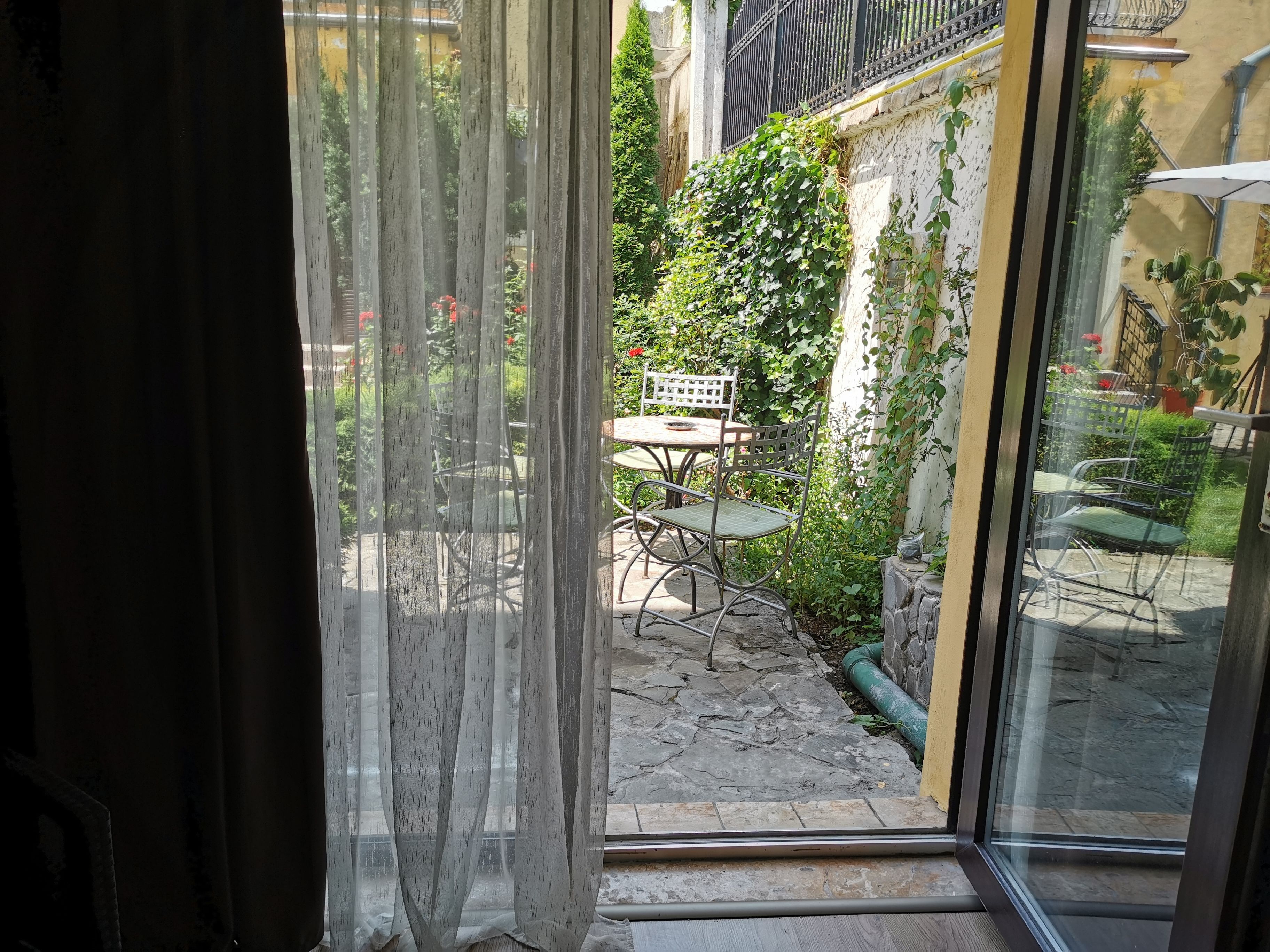 Visão através de uma porta de vidro para um pátio com uma mesa e cadeiras de metal, rodeado por vegetação e uma parede de pedra.