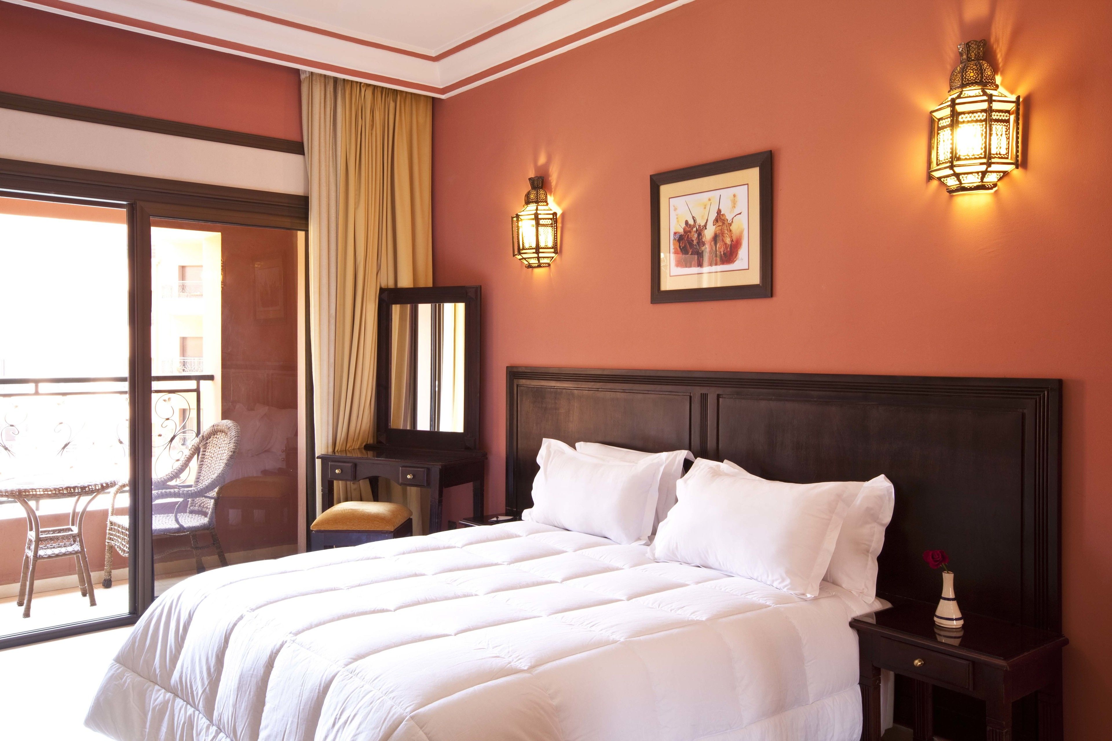 Quarto de hotel com uma cama grande, duas luzes fixadas na parede, um quadro emoldurado, uma pequena mesa com um vaso e uma cadeira perto de uma porta de vidro deslizante.