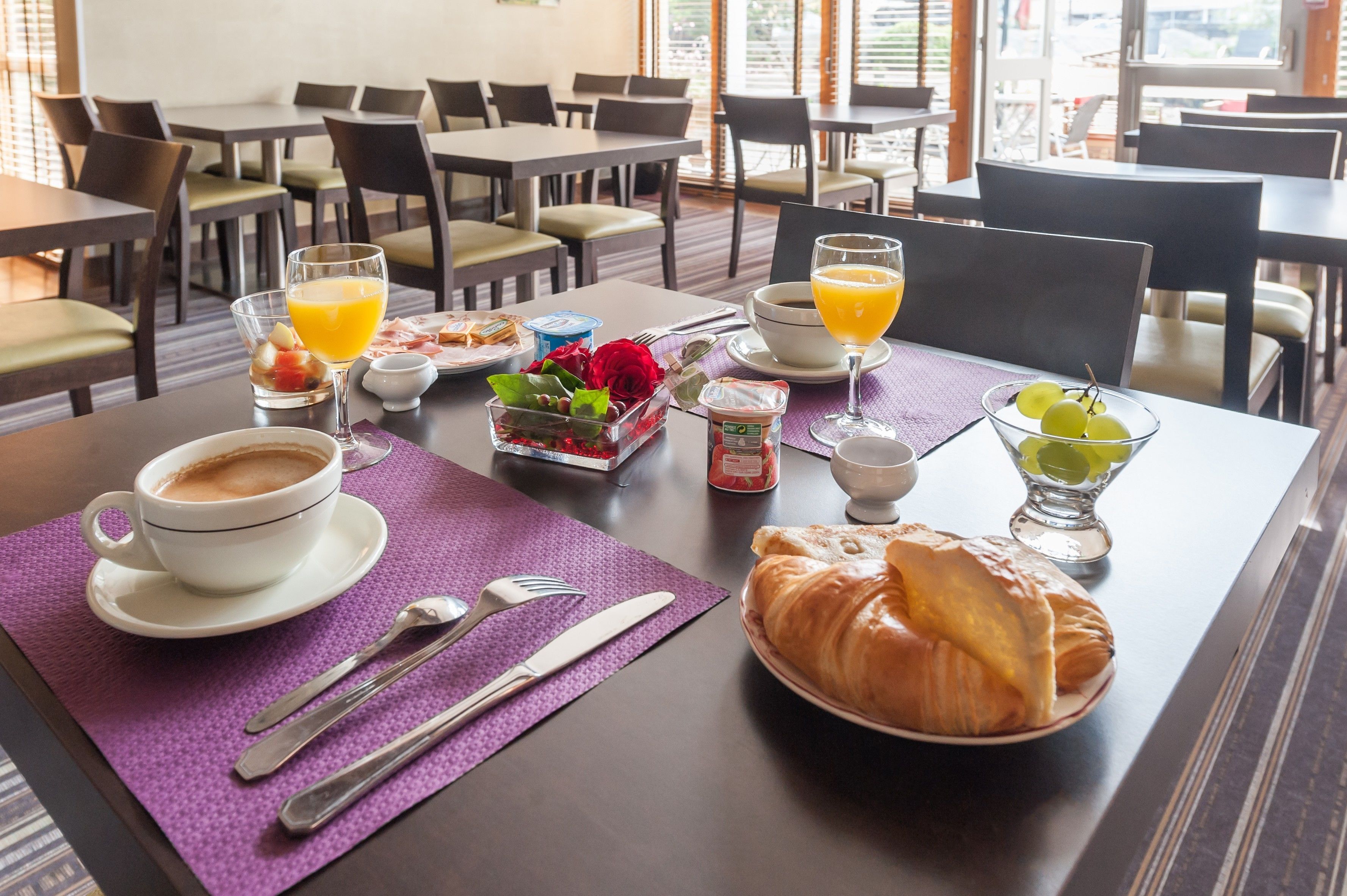 Mesa de café da manhã com um croissant, suco de laranja, café e uma cesta de frutas em uma área de refeições de hotel.